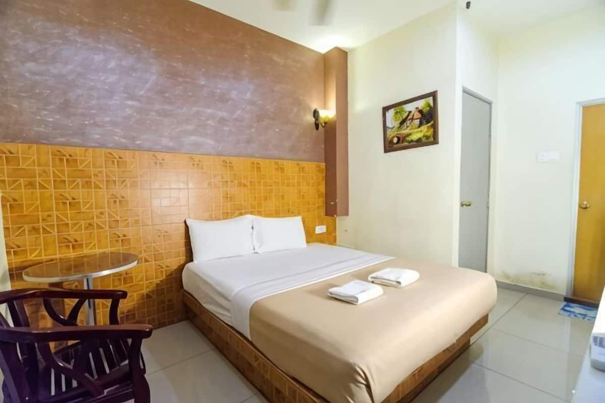 Quarto de hotel com uma cama, uma mesinha redonda, uma cadeira de madeira, uma luminária na parede e uma porta.