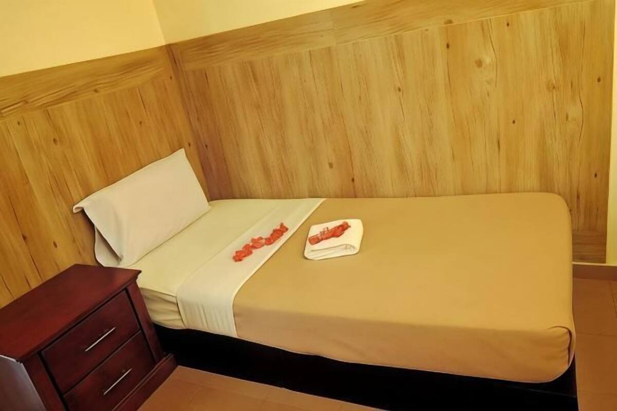 Uma cama de solteiro com cabeceira de madeira, travesseiro branco, colcha bege e toalha vermelha com estampa de coração.