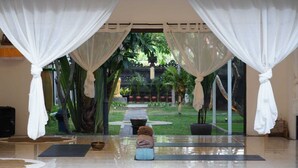 Um espaço interior tranquilo com vista para um jardim exuberante, com área de meditação, tapetes e uma tigela.