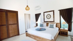 Quarto com uma cama grande, cabeceira de madeira e almofada decorativa. Há uma janela com cortinas, um ar condicionado fixado na parede e um lustre.