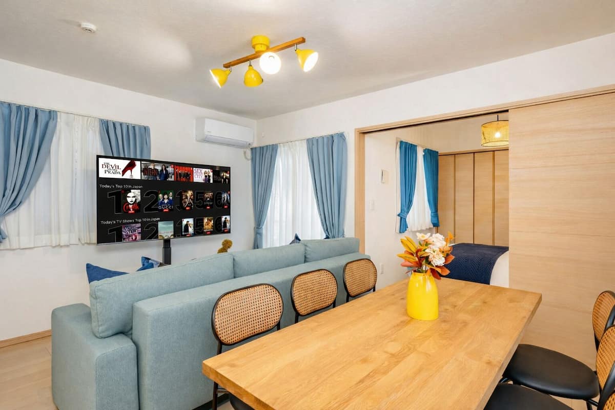Uma sala de estar moderna com sofá, mesa de jantar e cadeiras. Há uma TV na parede exibindo programas de televisão.