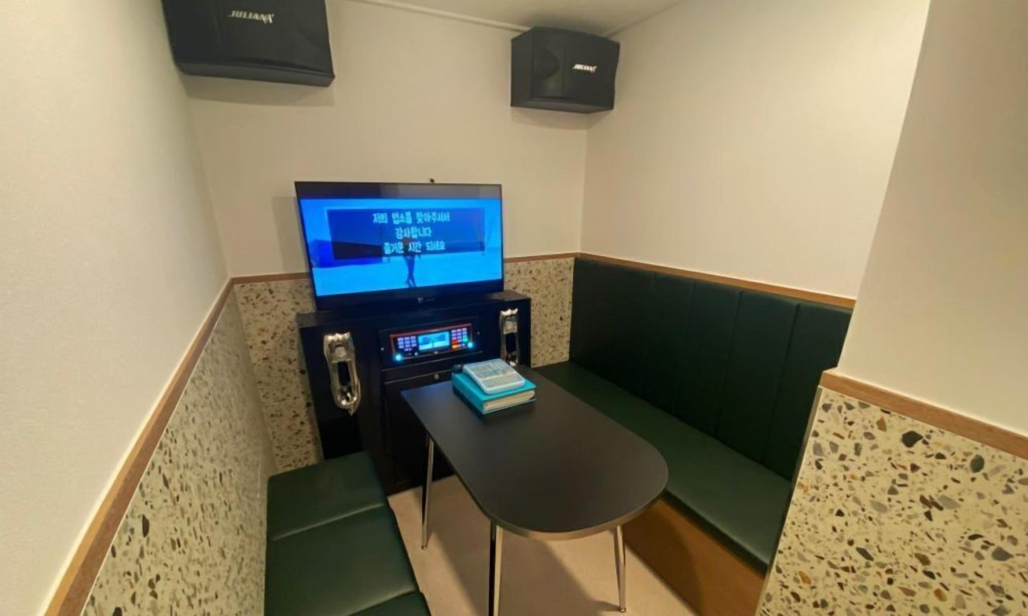 Um quarto pequeno e reservado com televisão, uma mesinha e um banquinho verde estofado.