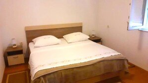 Uma cama de casal com cabeceira de madeira, duas almofadas brancas, uma mesa de cabeceira com abajur e uma gaveta pequena.
