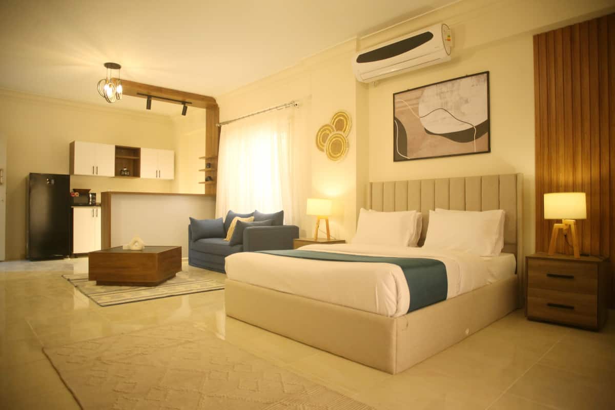 Quarto de hotel com cama, sofá, mesa de centro, escrivaninha e cozinha compacta.