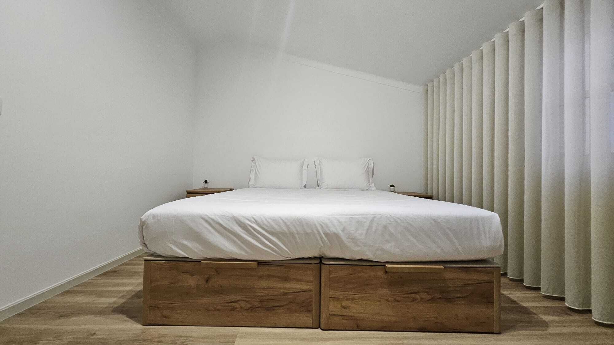 Quarto com cama ao centro, mesas de cabeceira de cada lado e janela com persianas verticais.