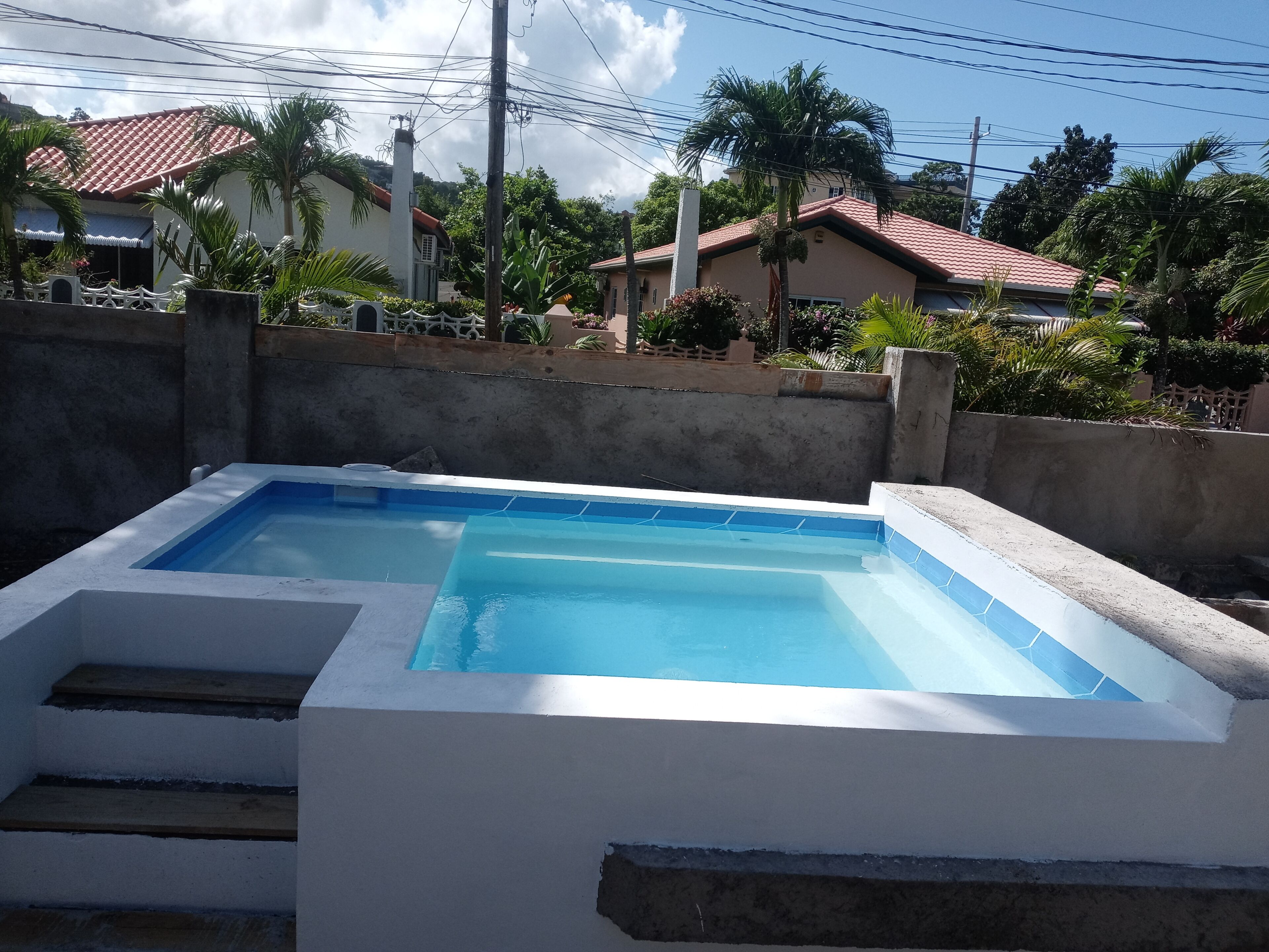 Uma piscina retangular com moldura de azulejos brancos e azuis, rodeada por um muro de concreto e degraus que levam à piscina.