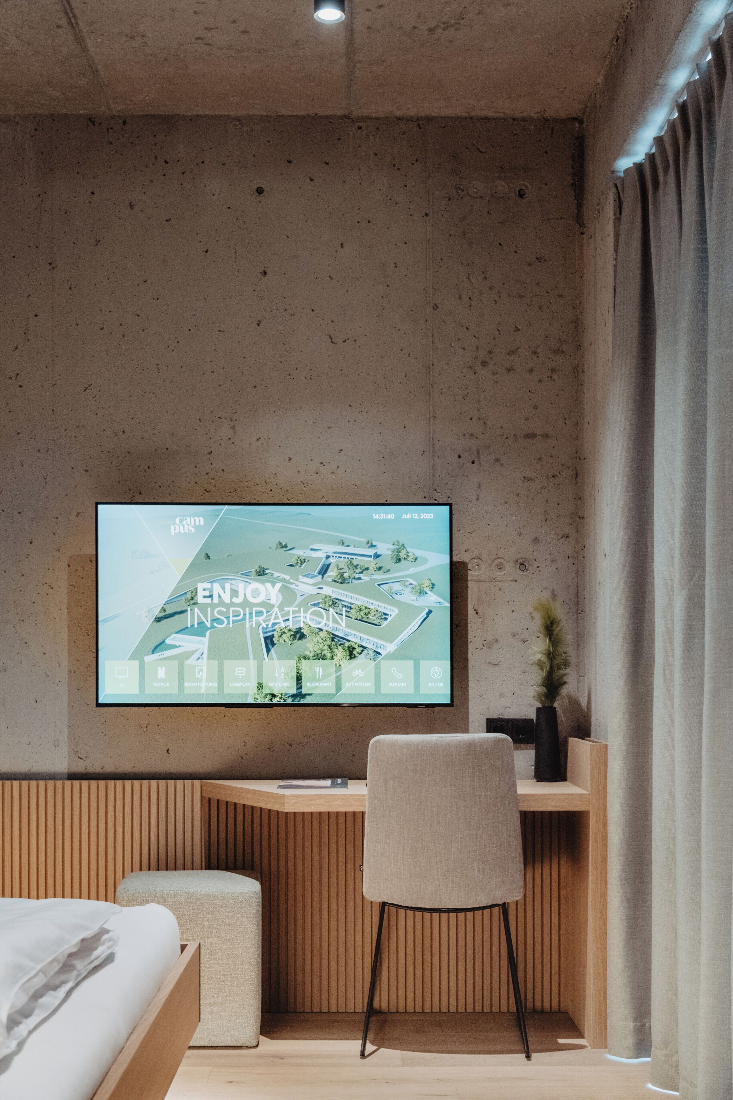 Quarto de hotel moderno com uma televisão de tela plana grande fixada em uma parede texturizada, uma escrivaninha de madeira com cadeira, uma cama com edredom branco e uma pequena planta em vaso.