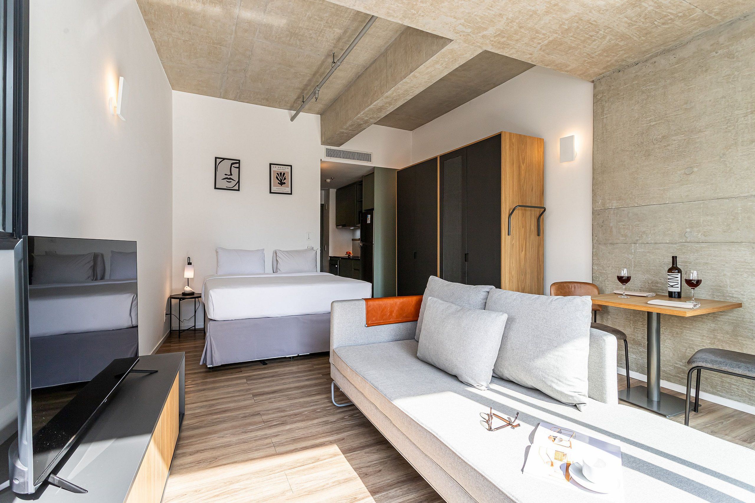 Quarto de hotel moderno com cama, sofá, televisão, mesa de jantar e cozinha compacta.