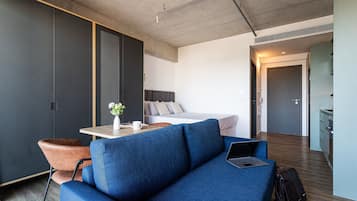 Quarto de hotel moderno com sofá azul, mesa de jantar de madeira, cama e cozinha compacta.