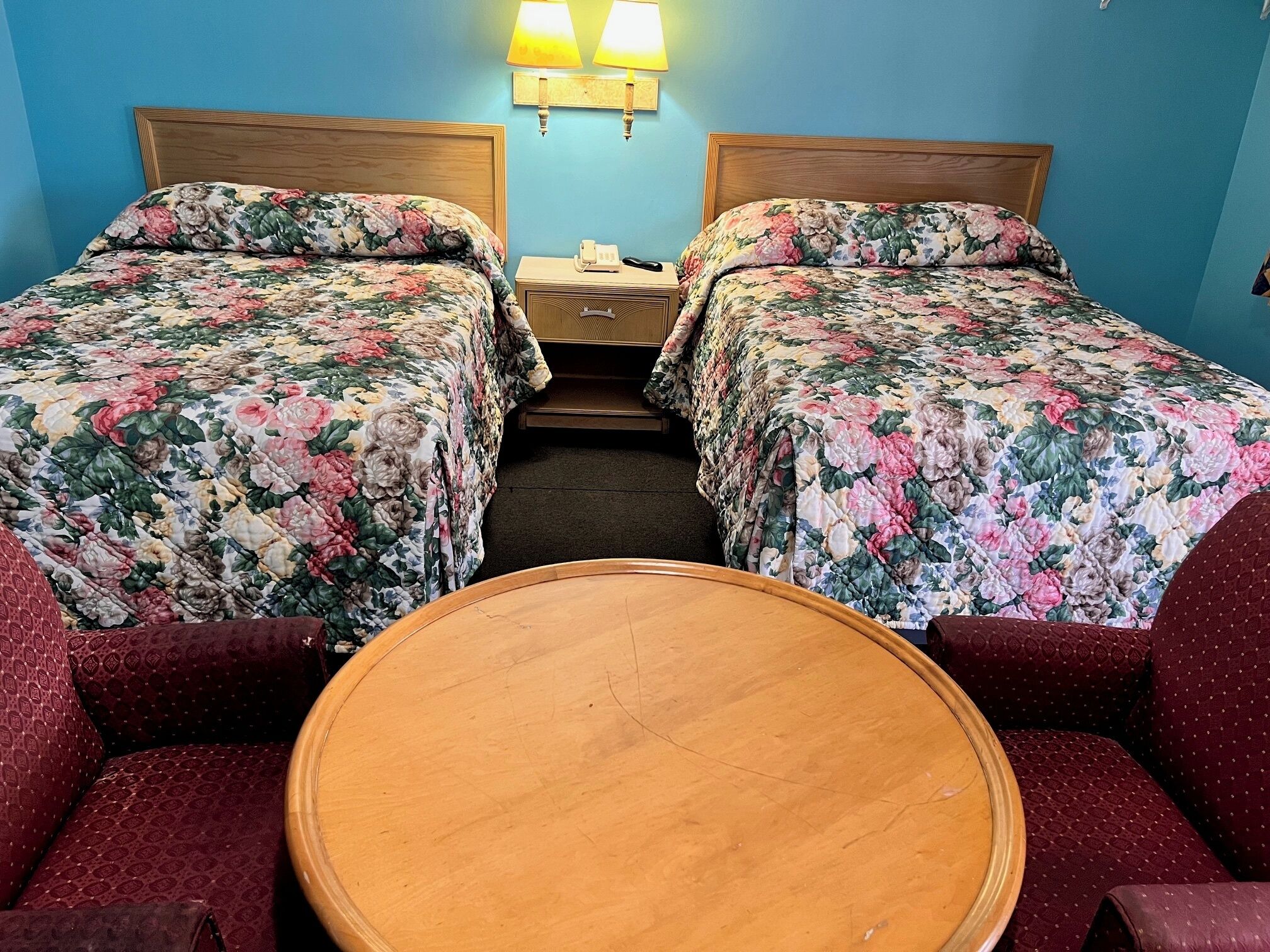 Quarto de hotel com duas camas, cabeceira de madeira, uma mesa pequena e duas luminárias fixadas na parede.
