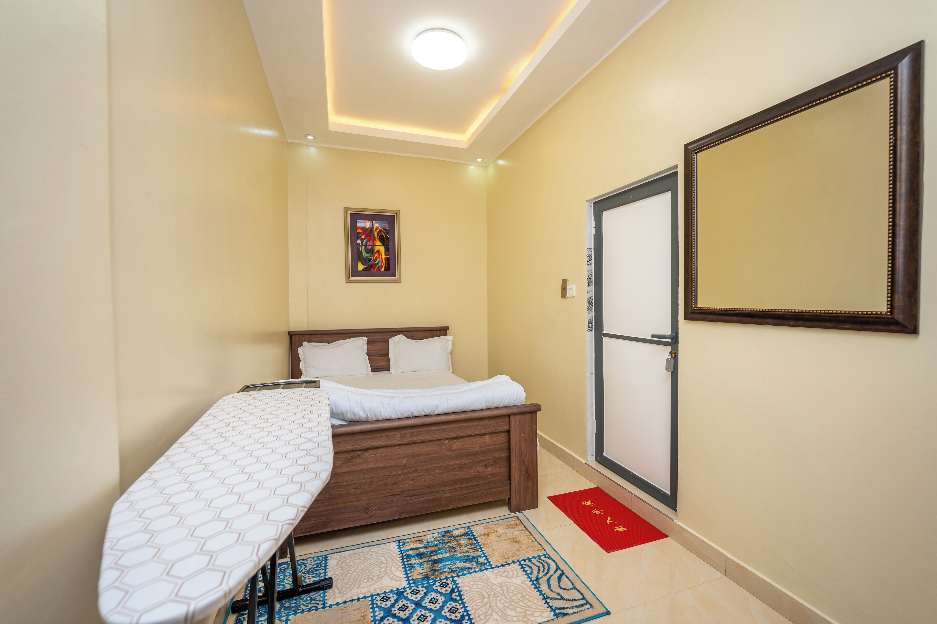 Um quarto com cama, tábua de passar roupa, tapete, espelho e porta.
