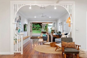 Uma sala ampla com piso de madeira, um tapete redondo, um sofá e uma poltrona. Há uma escada com corrimão branco que leva a uma varanda.