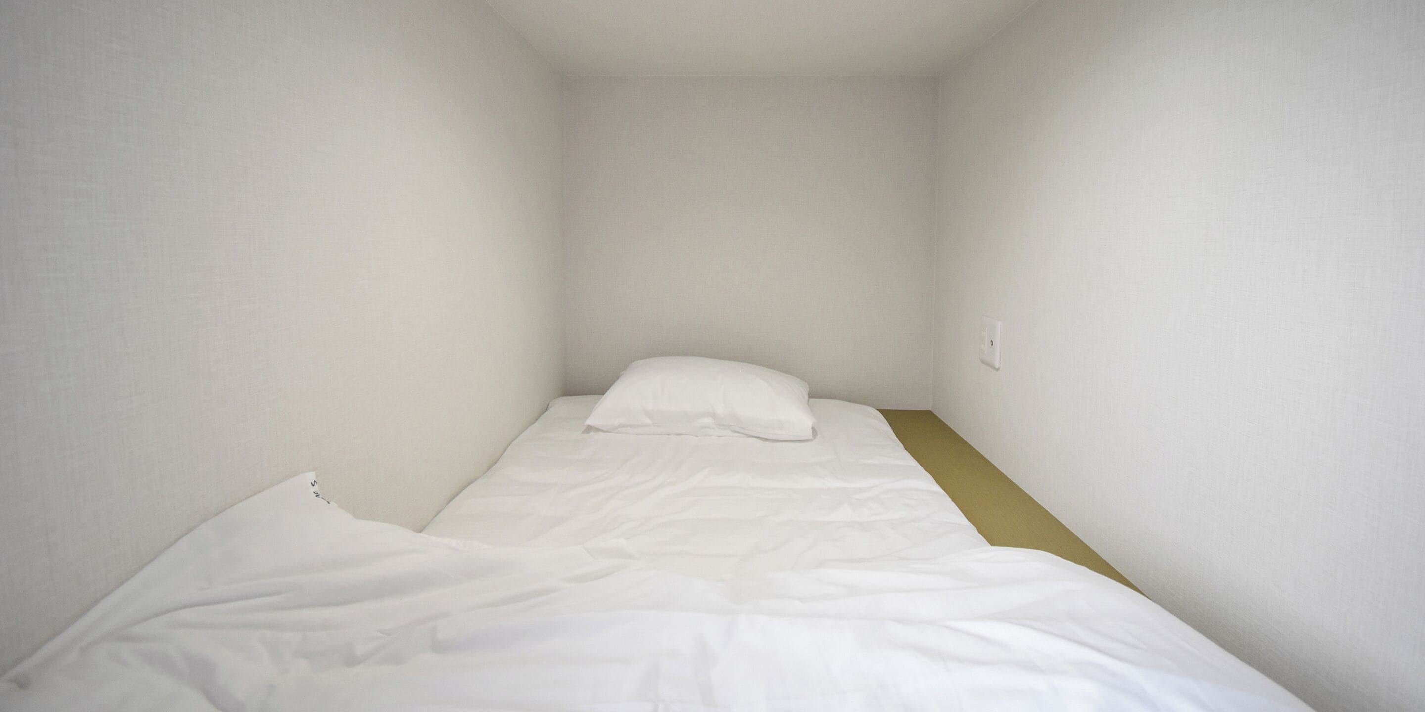 Quarto pequeno com uma cama de solteiro, roupa de cama branca e chão de madeira.