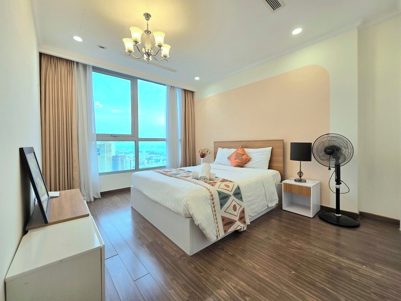 Quarto de hotel moderno com uma cama grande, um ventilador e vista para a cidade pela janela.