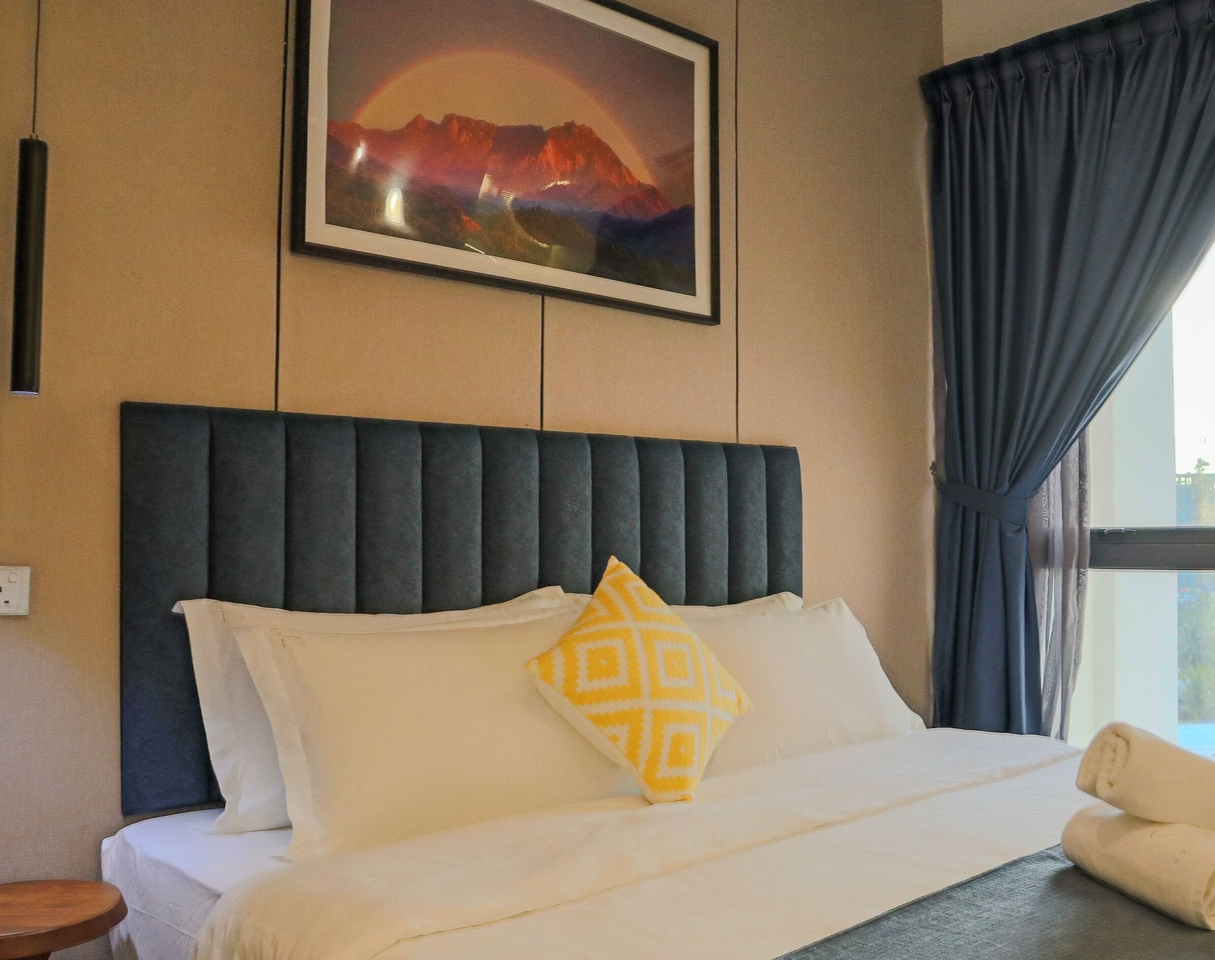 Quarto de hotel com uma cama grande, cabeceira, pintura de uma paisagem montanhosa, janela com cortinas e uma pequena mesa.