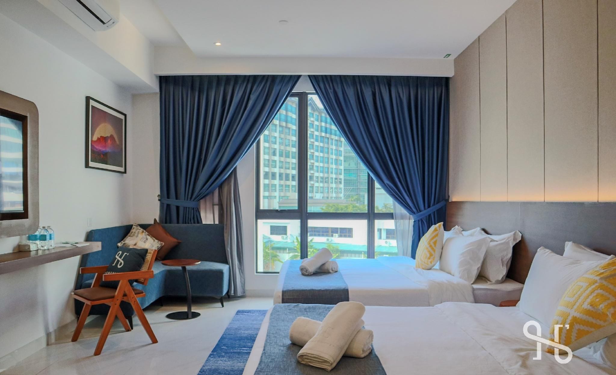Quarto de hotel com cama grande, sofá, escrivaninha e uma cadeira. Há uma janela com cortinas, um quadro na parede e vista para prédios.