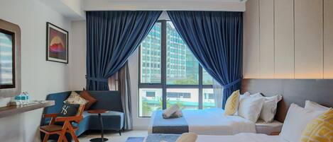 Quarto de hotel com cama grande, sofá, escrivaninha e uma cadeira. Há uma janela com cortinas, um quadro na parede e vista para prédios.