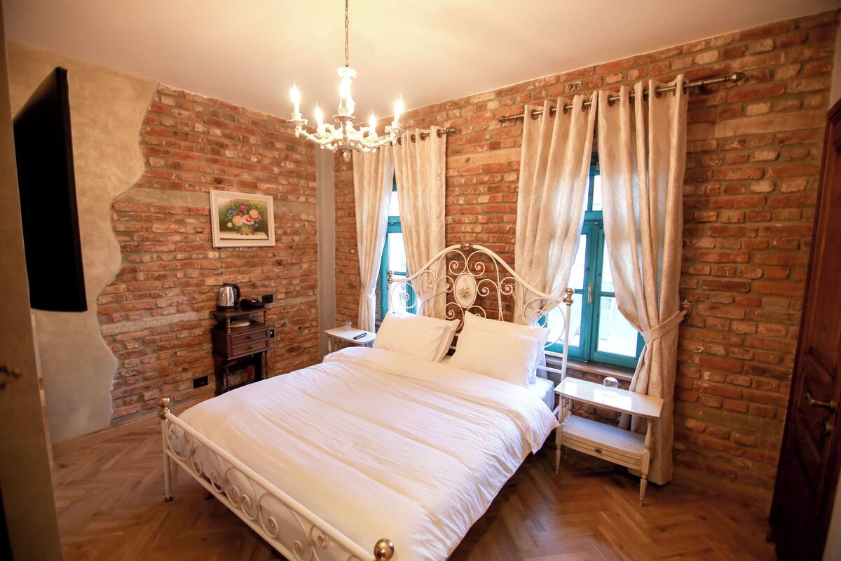 Quarto com parede de tijolos, cama branca, candelabro e janela com cortinas.