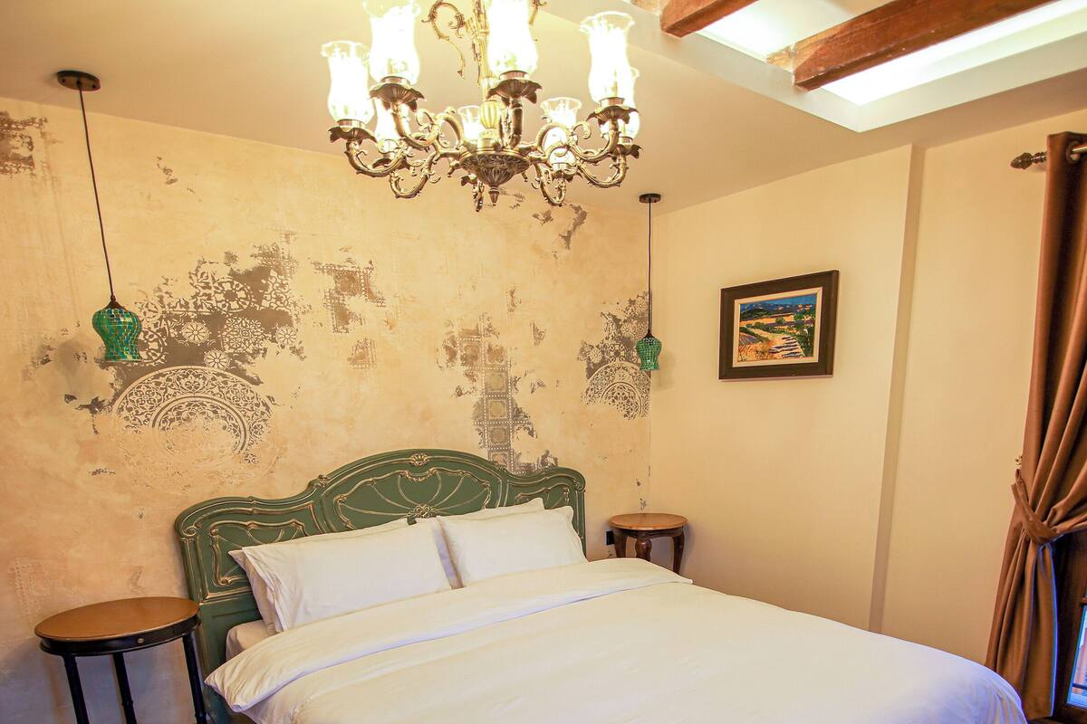 Um quarto com cama estofada verde, roupa de cama branca, um candelabro, uma mesa pequena e uma pintura na parede.