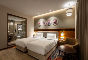 Quarto de hotel com duas camas, um espaço de estar com uma cadeira, uma mesa pequena, um espelho e um banheiro visível através da porta de vidro.