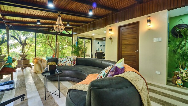 Uma sala de estar moderna com um sofá preto curvo, uma mesinha e um candelabro. Há vigas de madeira no teto e vista para árvores lá fora.