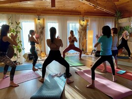 Um grupo de pessoas em uma aula de yoga, praticando em um espaço com piso de madeira e janelas amplas.