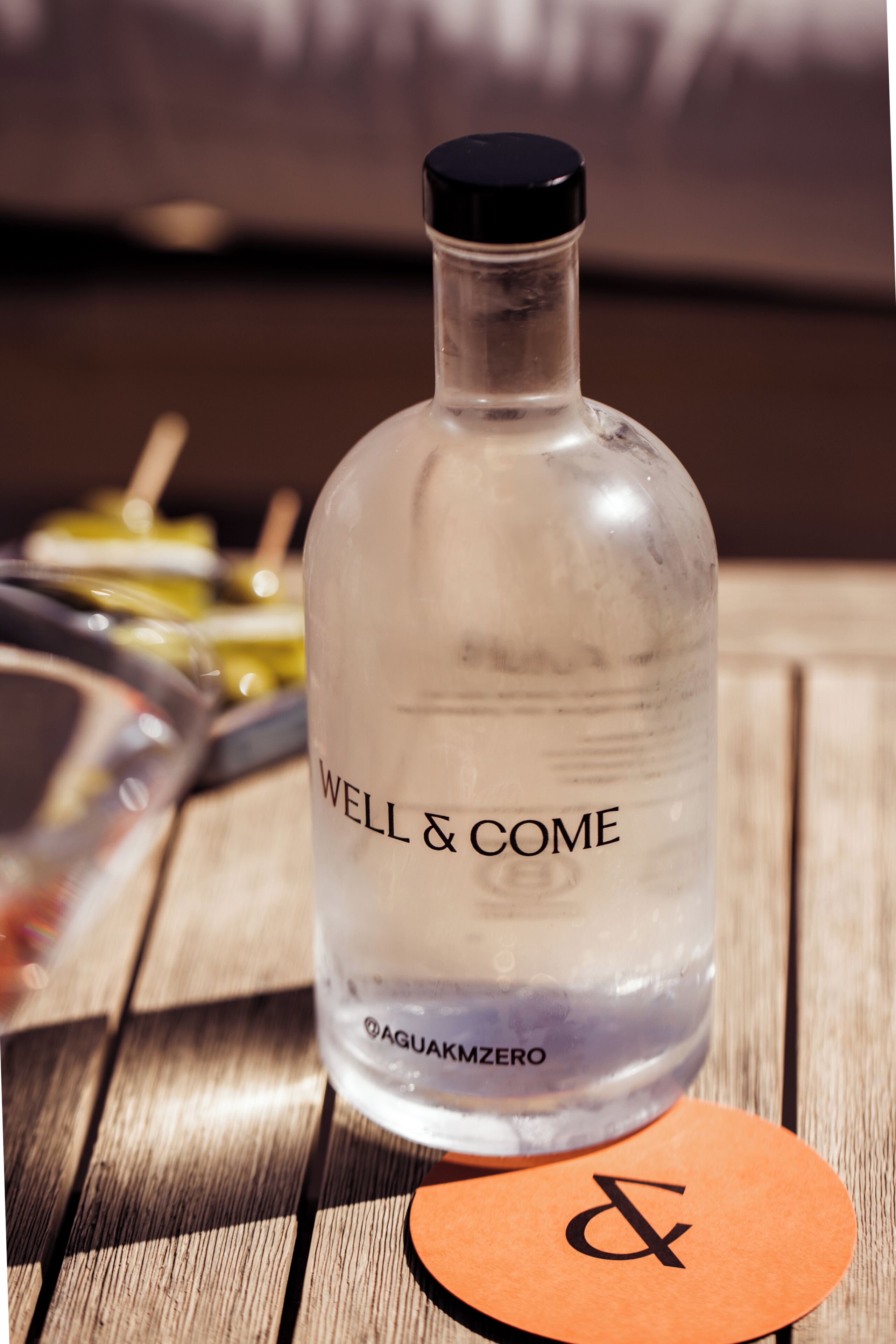 Garrafa de vidro transparente com o rótulo 'WELL & COME' sobre uma superfície de madeira, acompanhada de um apoio para copos e outro copo.