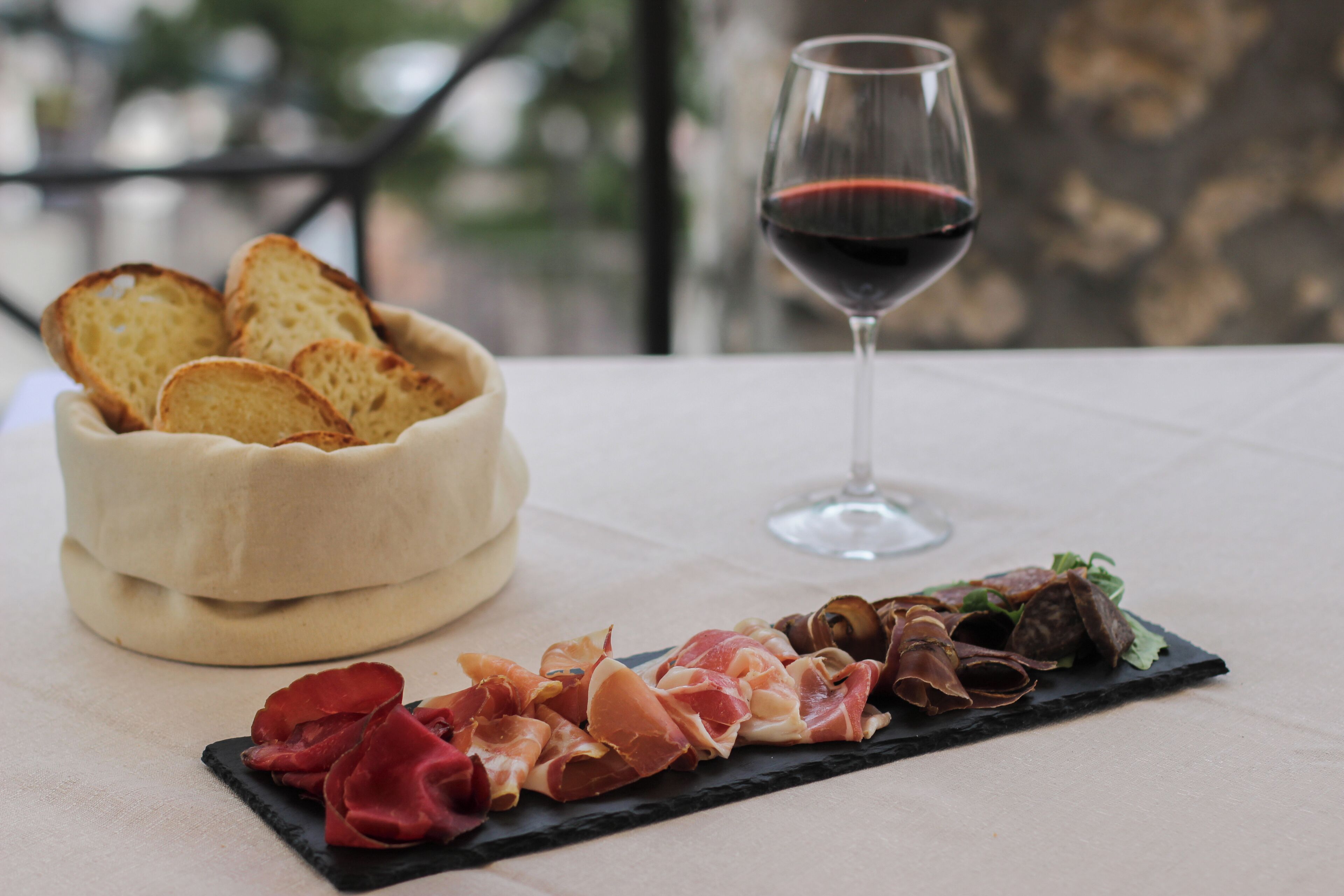 Uma mesa com uma tigela de pão, um copo de vinho tinto e um prato de embutidos.