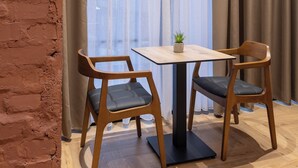 Quarto com parede de tijolos, uma mesa com duas cadeiras, uma planta pequena e uma janela com cortinas leves.