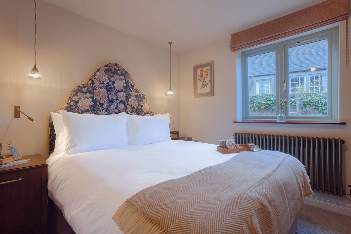 Uma cama bem arrumada com cabeceira florida, uma mesa de cabeceira com abajur e uma janela com vista para o verde.