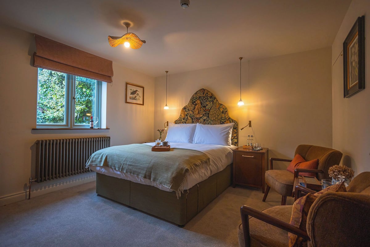 Quarto com uma cama grande, mesas de cabeceira, uma cadeira, radiador e janela com vista para árvores.
