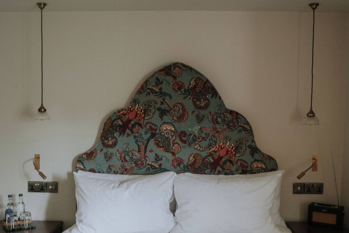 Uma cama com cabeceira estampada, duas mesas de cabeceira com abajures e um rádio fixado na parede.