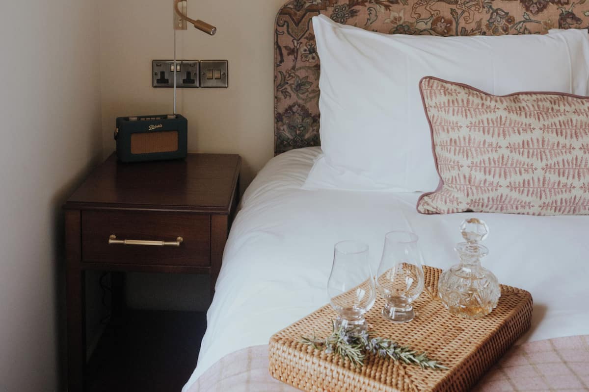 Cama bem arrumada com cabeceira estampada, mesa de cabeceira com rádio e bandeja com copos e garrafa.