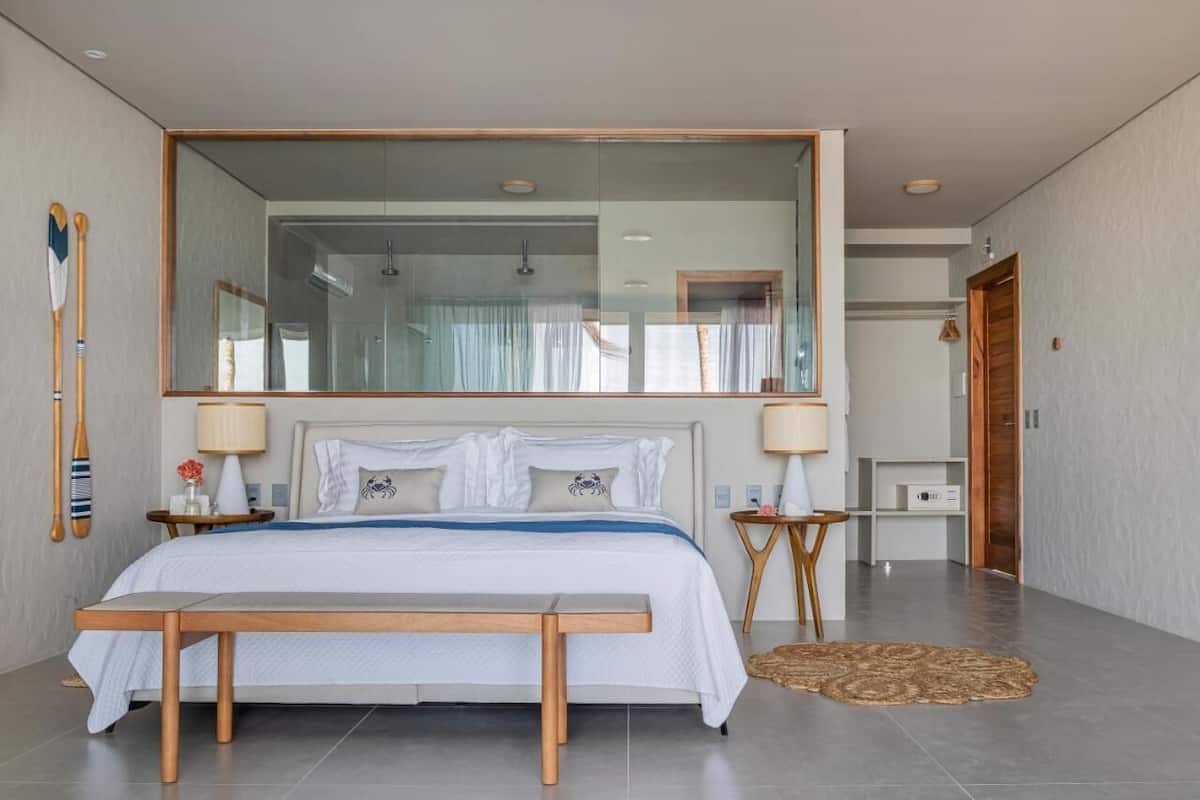 Quarto moderno com cama grande, mesas de cabeceira, um banco e um espelho amplo.