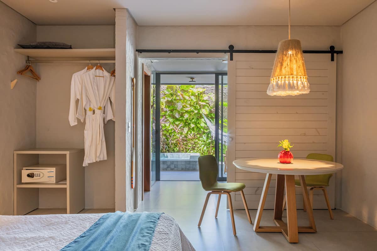 Quarto moderno com cama, mesa de cabeceira e um pequeno armário. Há uma janela com vista para o verde lá fora.