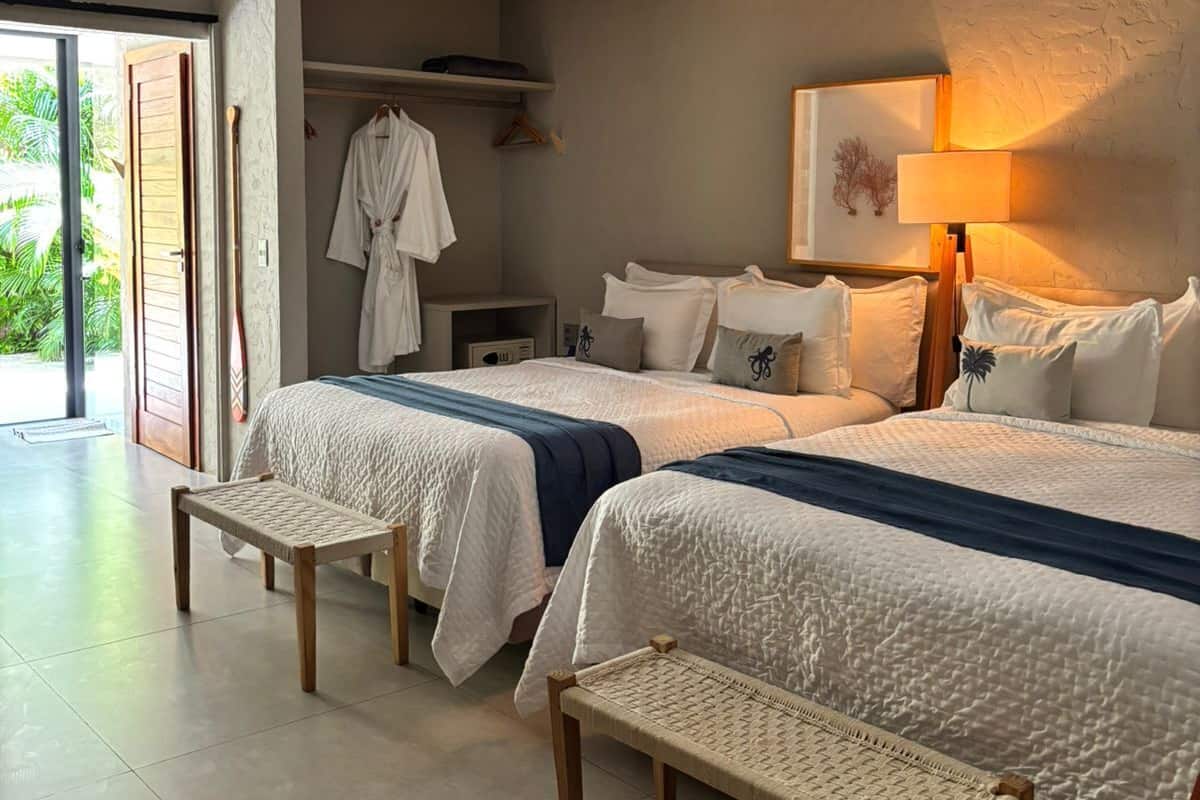 Quarto de hotel com duas camas, um banco, roupão e luminária.