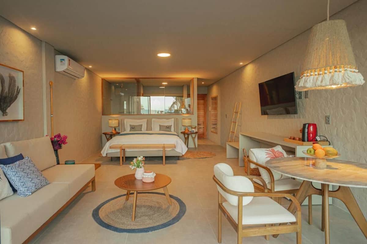 Quarto de hotel moderno com cama, sofá, mesa de jantar e televisão fixada na parede.