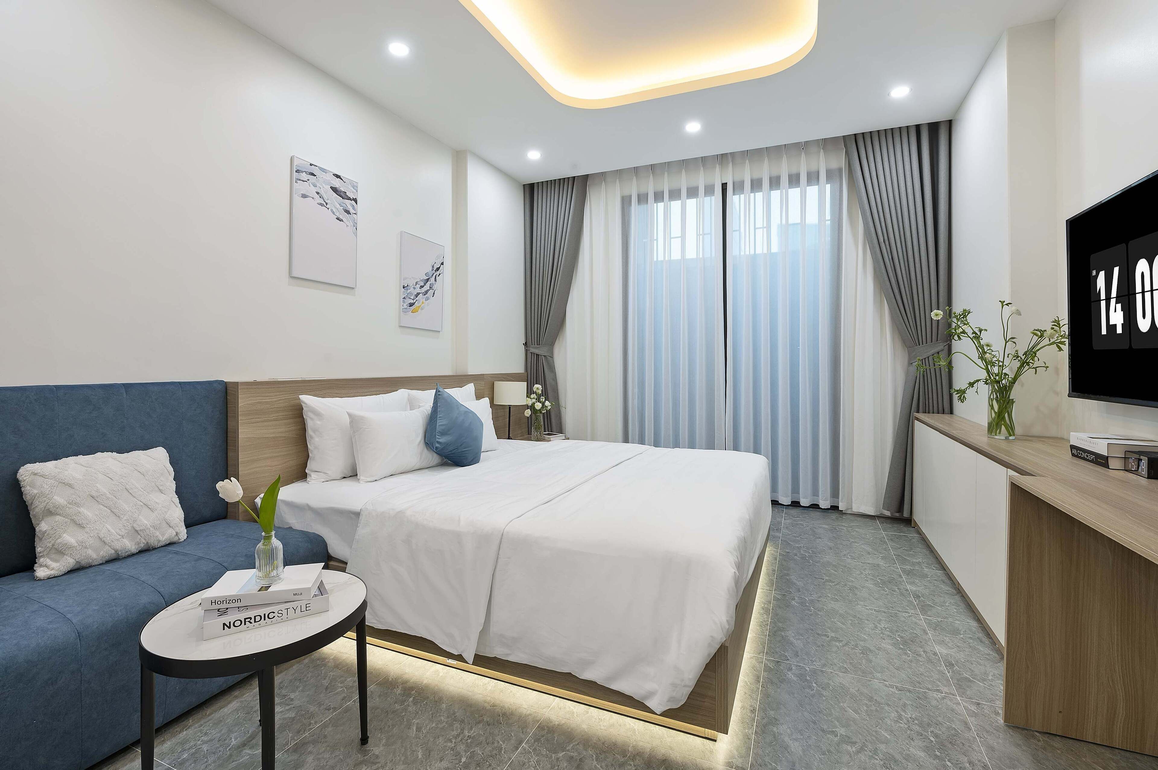 Quarto de hotel moderno com cama, sofá, mesinha com livros, televisão e janela com cortinas.
