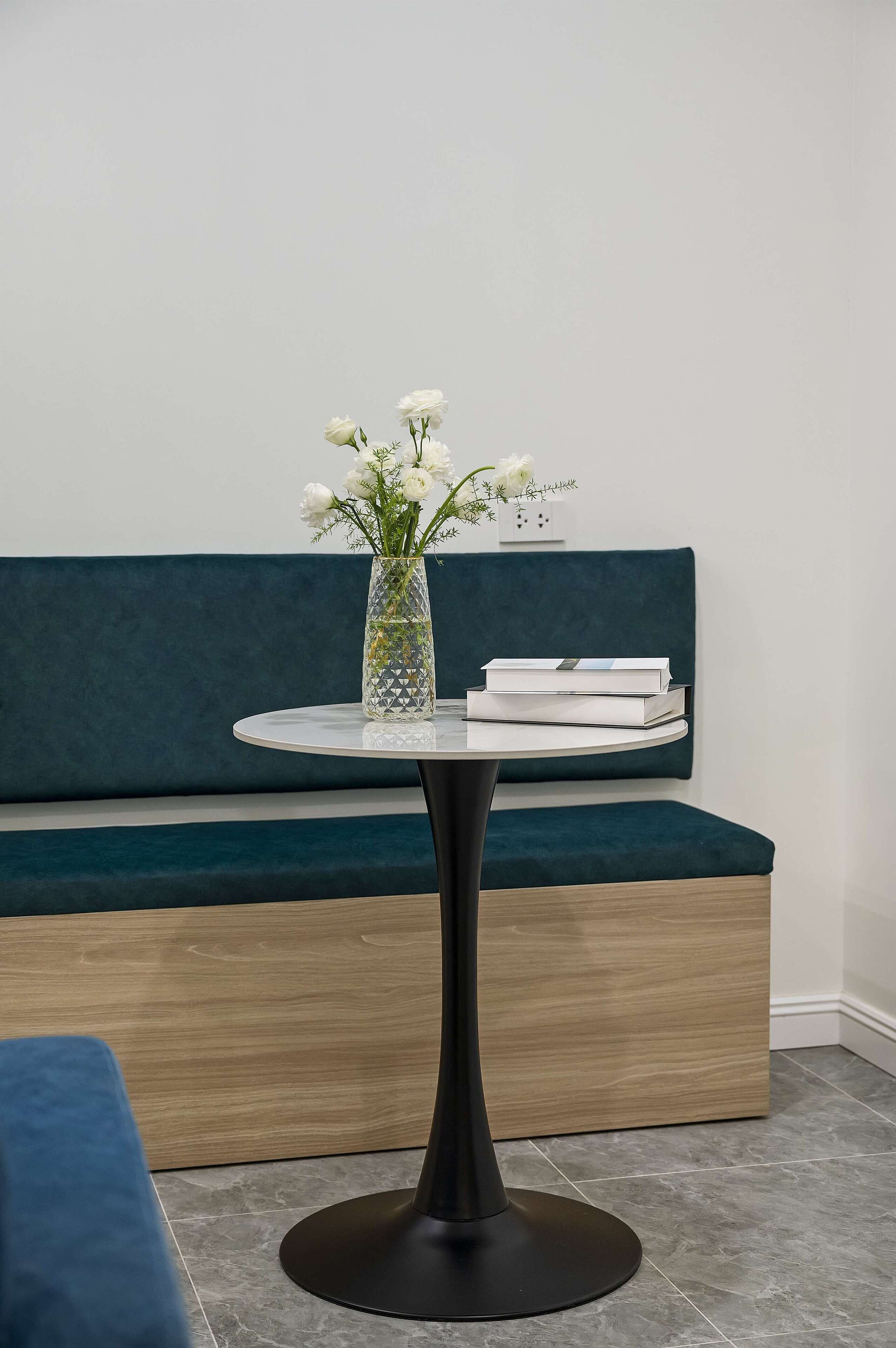Uma mesa redonda com um vaso de flores brancas, uma pilha de livros e um banco estofado azul ao fundo.