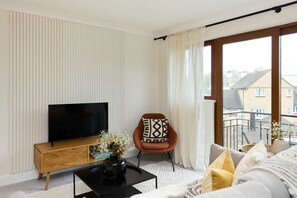 Uma sala de estar moderna com uma televisão de tela plana, um móvel de madeira para TV, uma poltrona marrom com almofada estampada, um sofá com almofadas e uma janela grande com vista para edifícios.