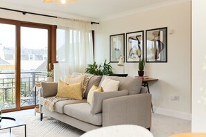 Uma sala de estar moderna com um sofá, uma mesa de centro e vista para o exterior. Há duas molduras na parede e uma planta sobre uma mesinha lateral.