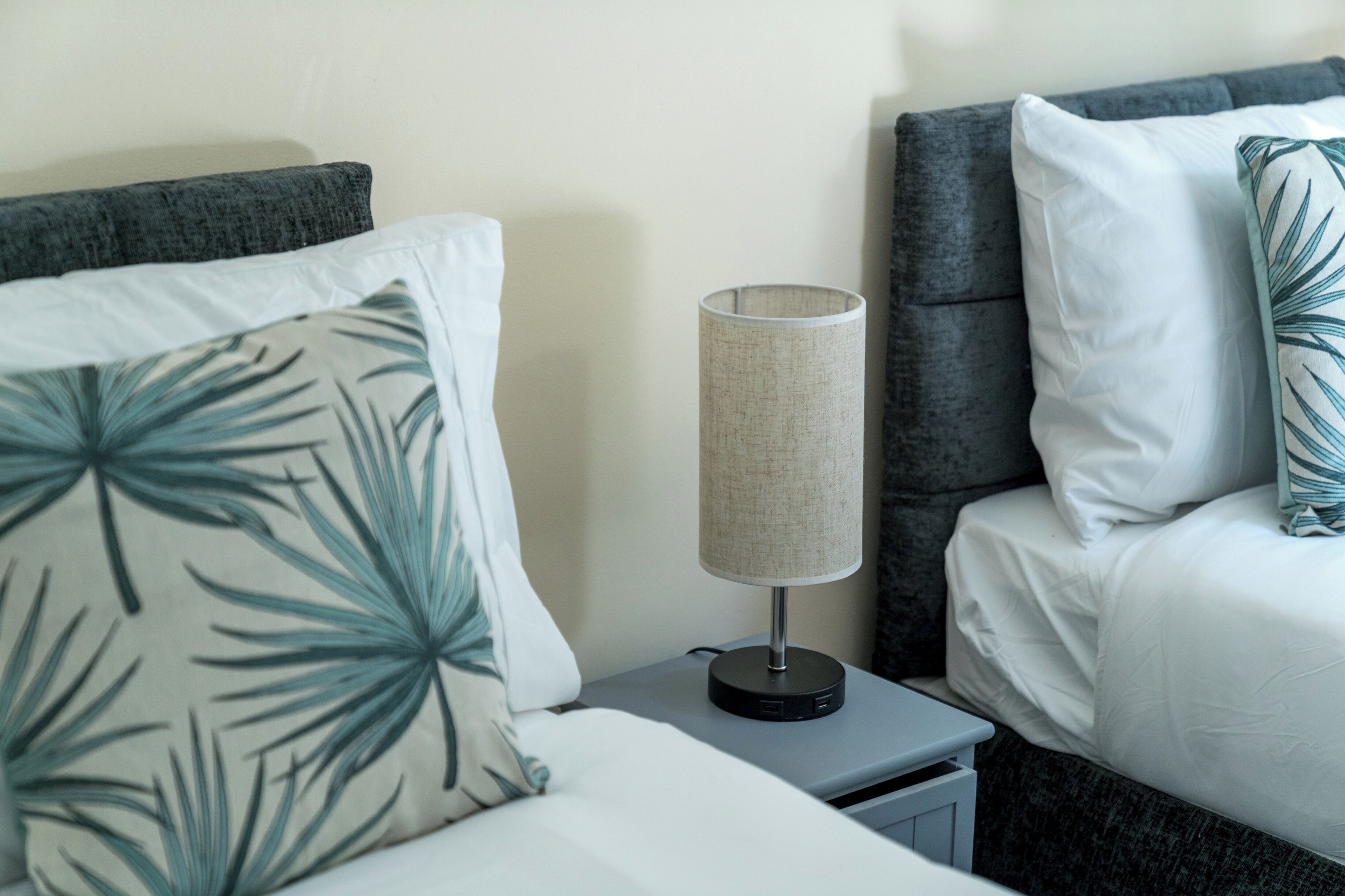 Uma cama bem arrumada com travesseiro branco e azul-esverdeado estampado com folhas, cabeceira cinza e mesa de cabeceira com abajur.
