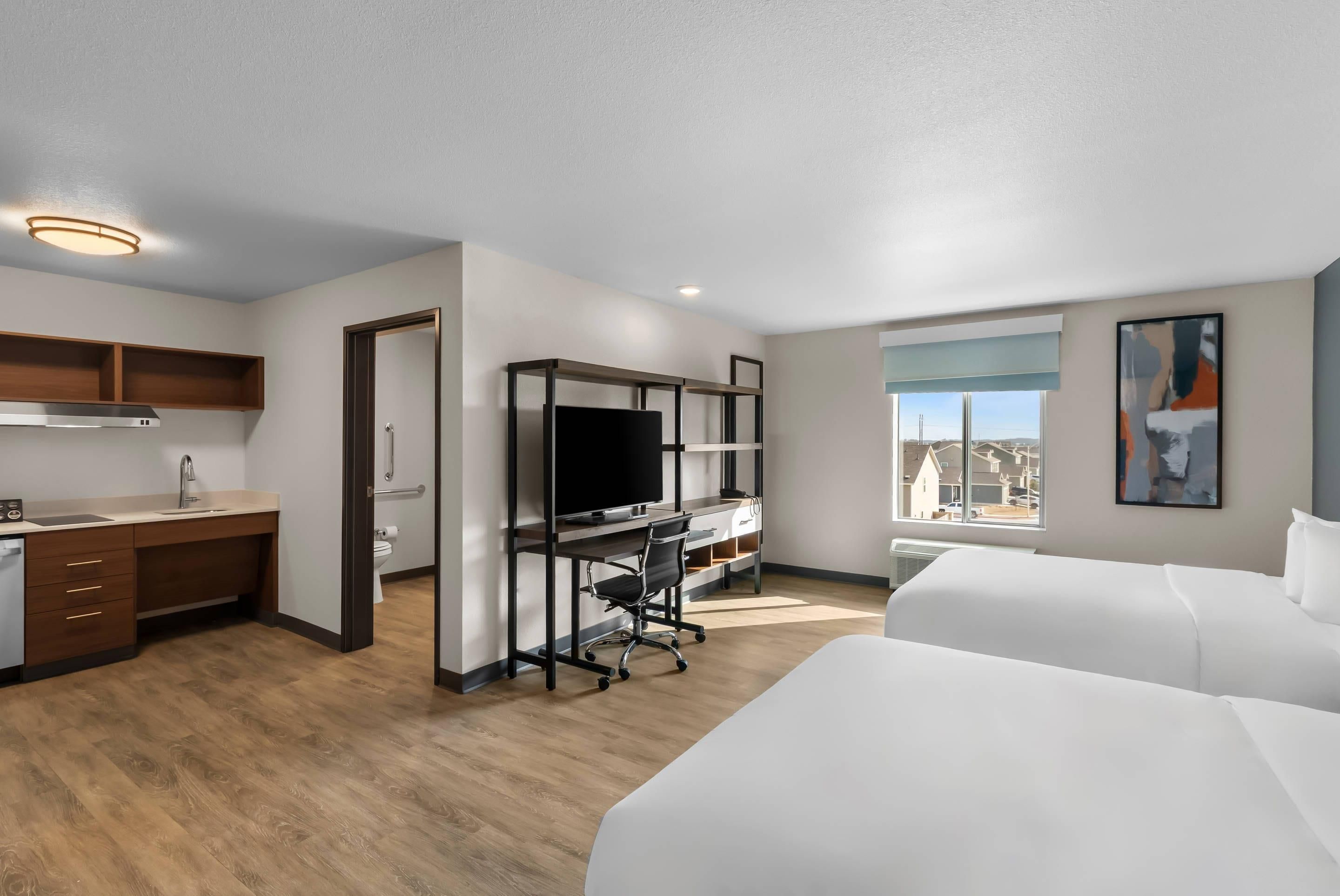 Quarto de hotel moderno com piso de madeira, uma escrivaninha com computador, televisão e uma cama.