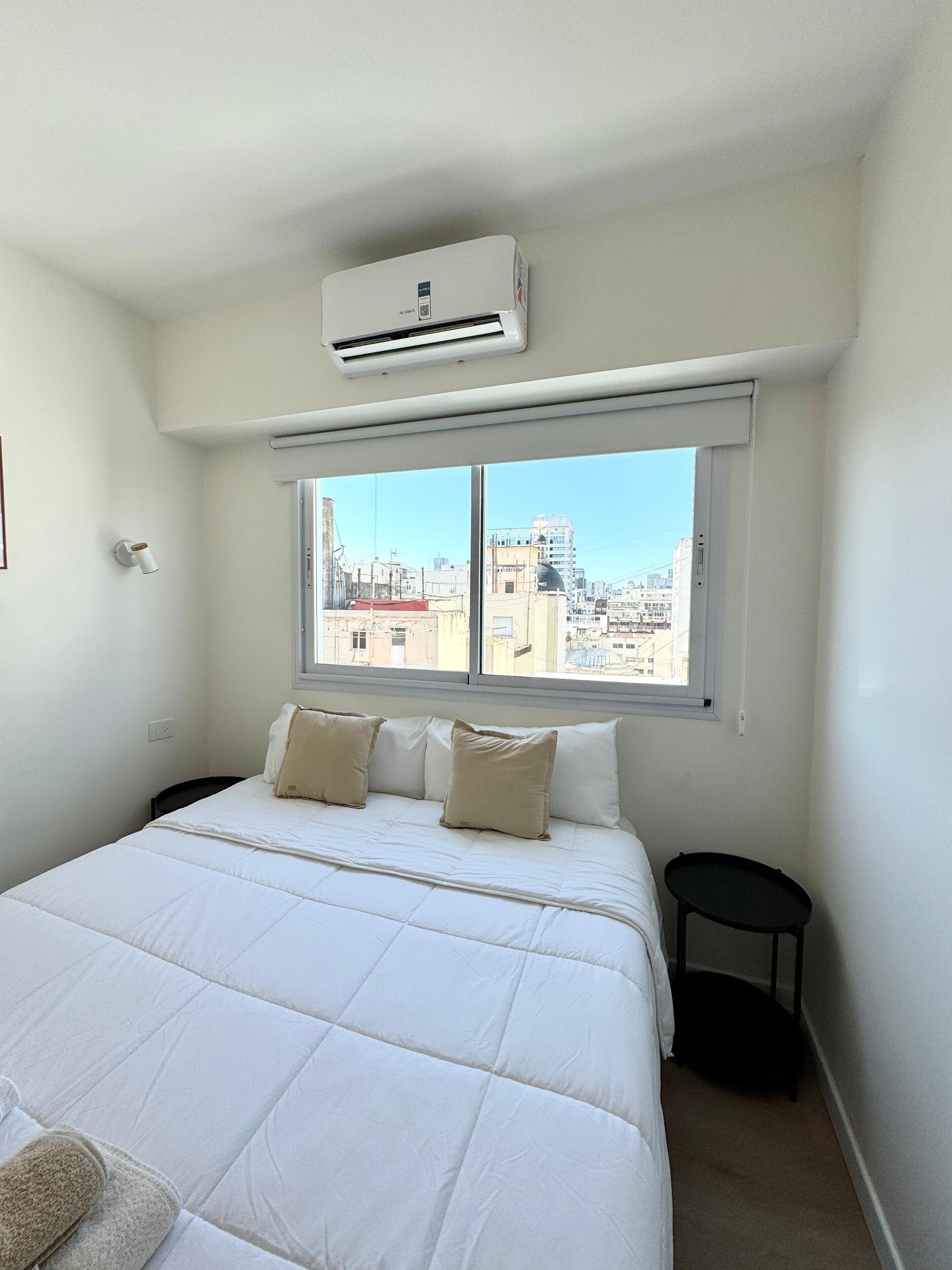 Quarto com cama grande, janela com vista para a cidade, ar condicionado e uma pequena mesa.