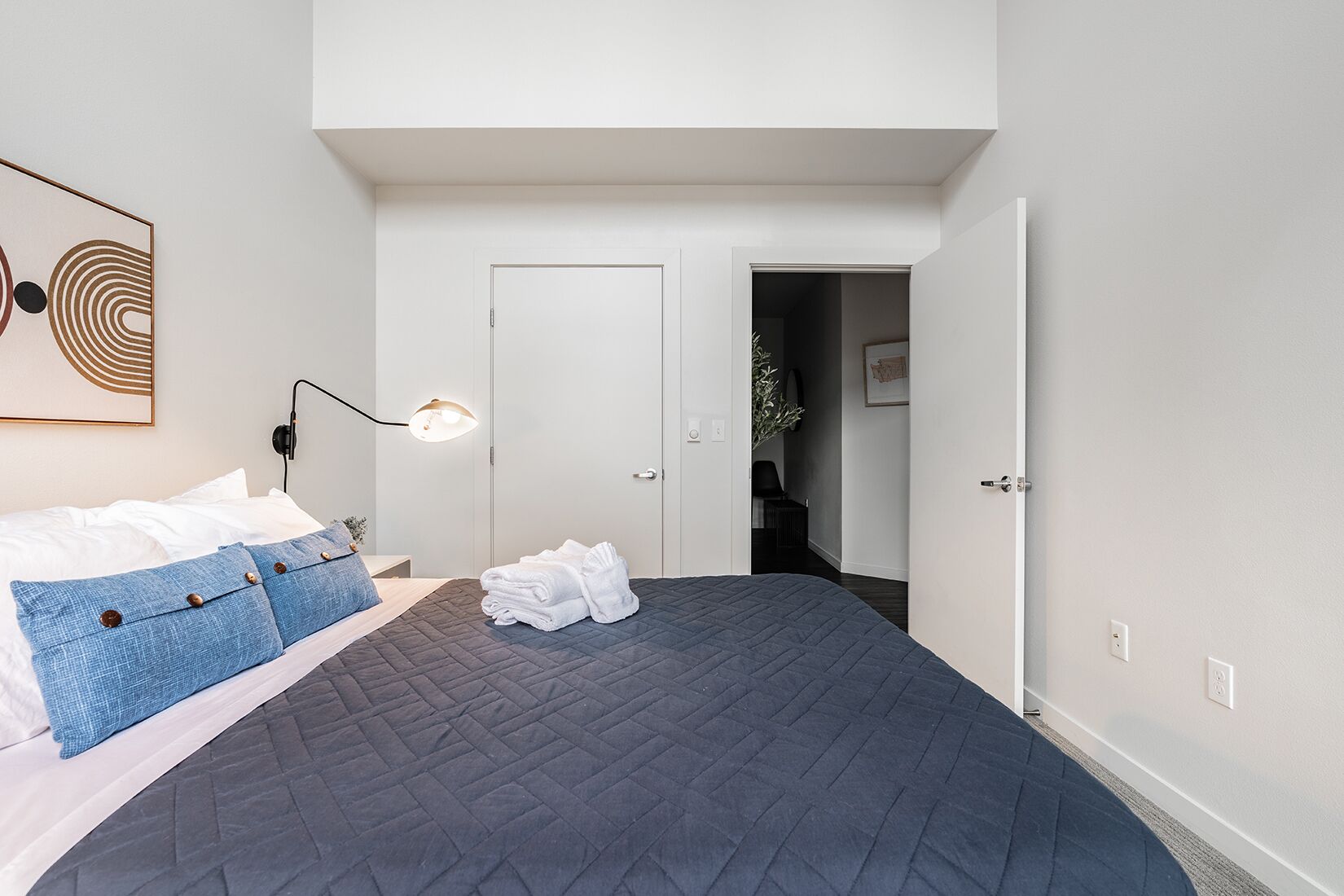 Quarto moderno com uma cama grande, uma mesinha de cabeceira, uma luminária na parede e uma porta que dá para outro espaço.