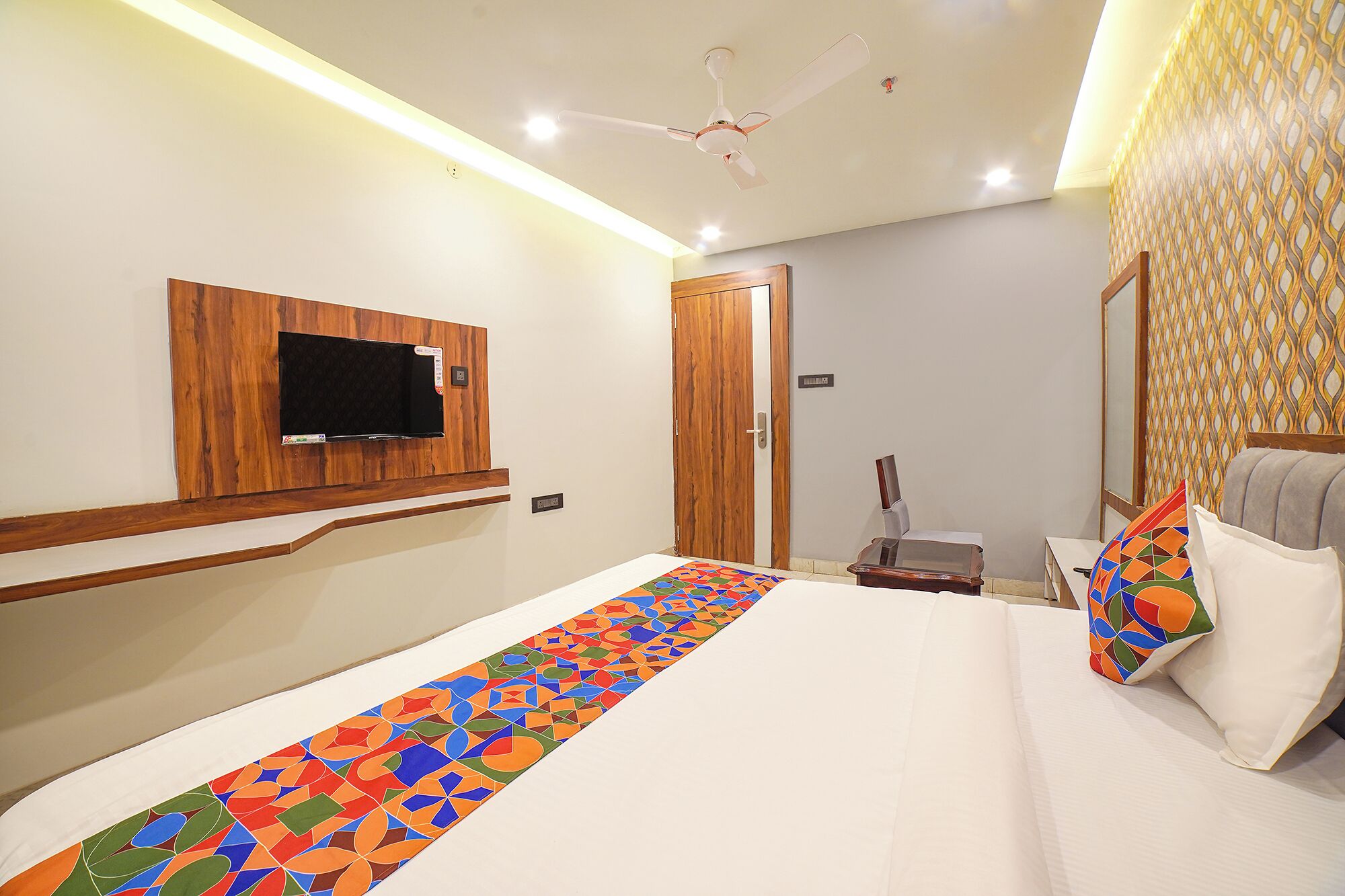 Quarto de hotel com uma cama grande, televisão fixada em um painel de madeira, ventilador de teto e carpete estampado.
