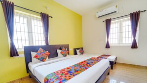 Quarto de hotel com uma cama grande, duas janelas com cortinas, um aparelho de ar condicionado e piso ladrilhado.