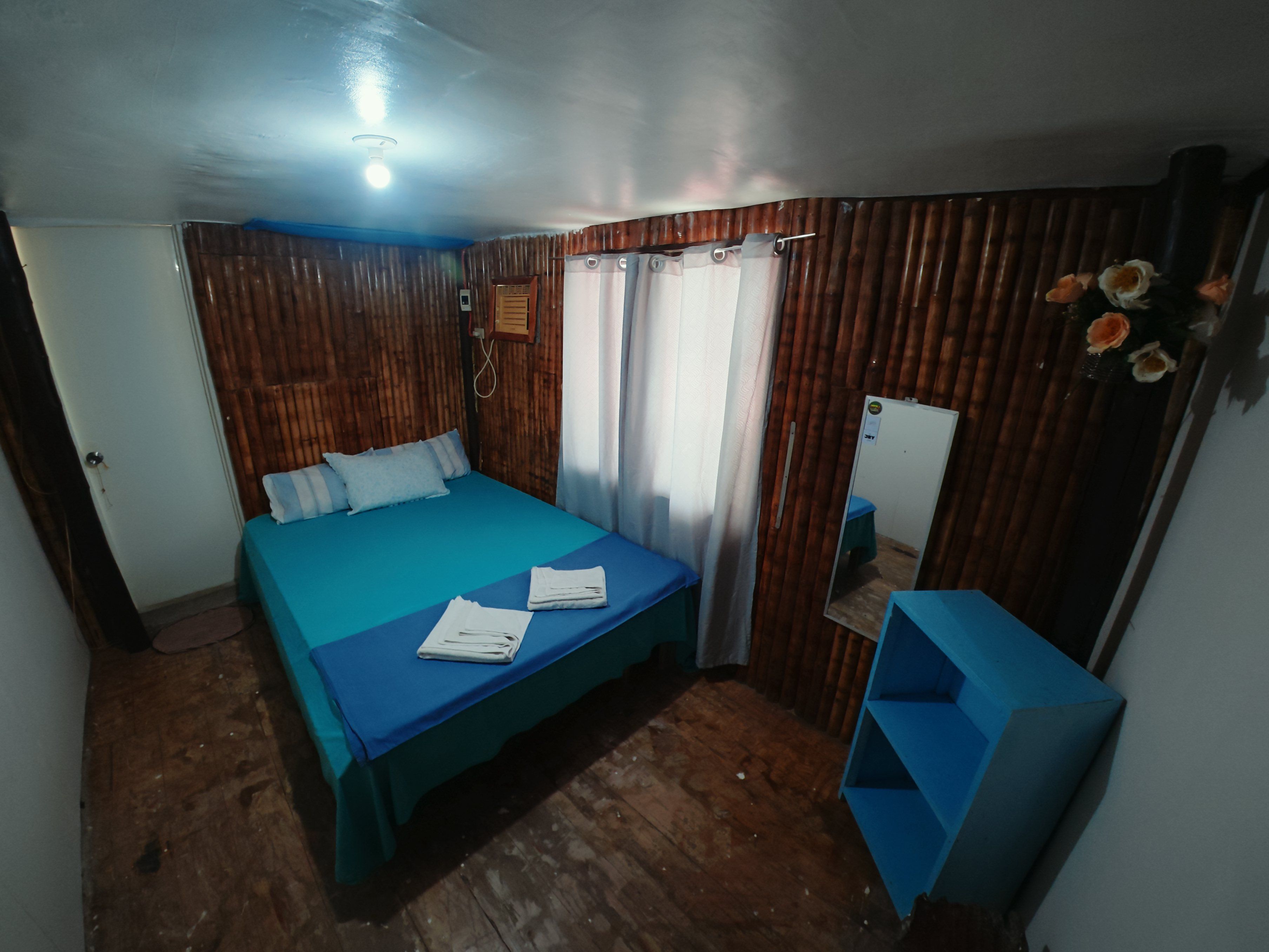 Quarto com cama, escrivaninha pequena, espelho e janela com cortinas.