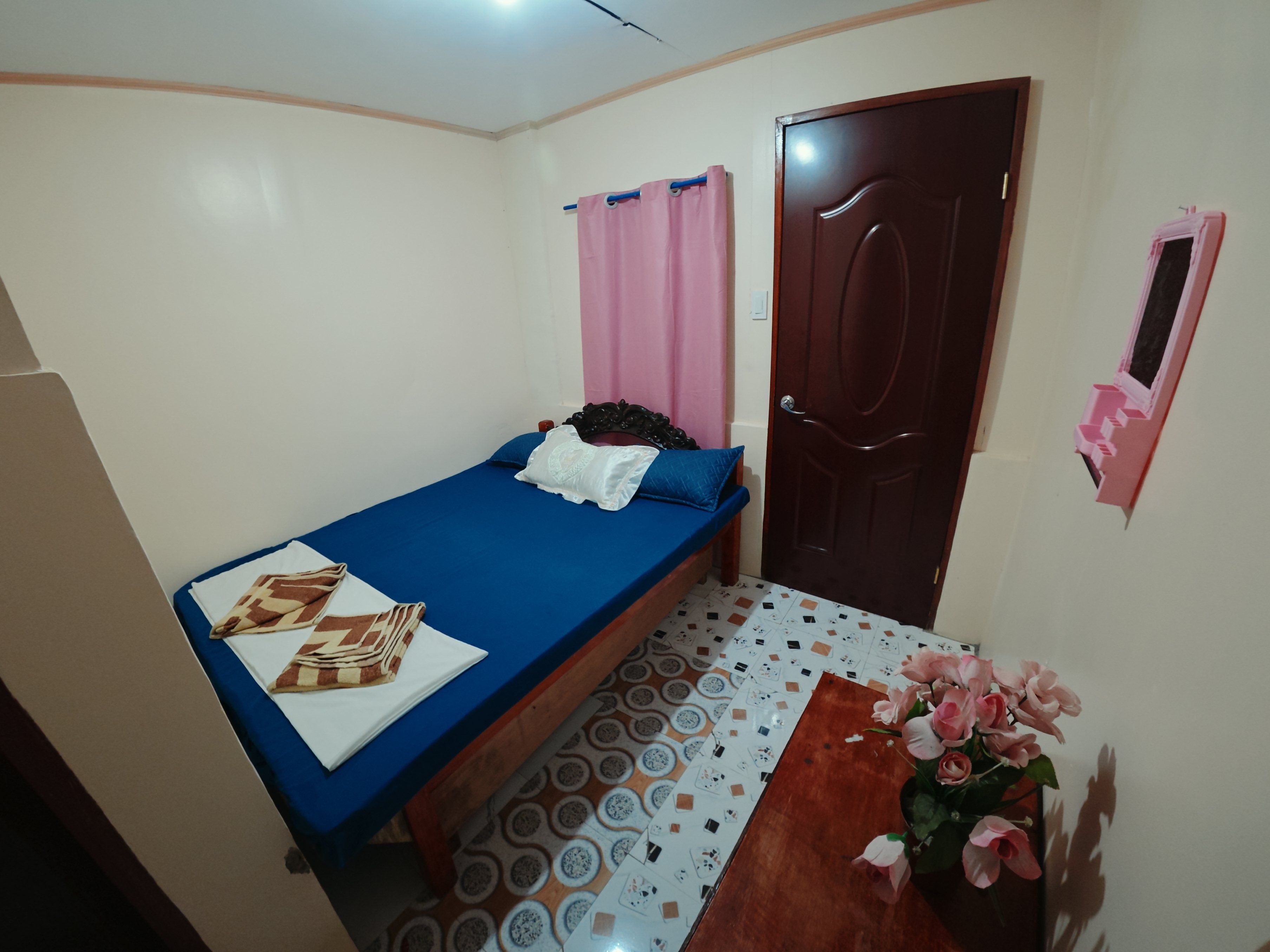 Quarto com uma cama de solteiro, porta de madeira, mesa pequena com um vaso de flores e chão estampado.