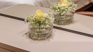 Cama bem arrumada com lençóis e almofadas brancas, mesa de cabeceira com dois vasos de vidro contendo flores, e um espelho refletindo a cama e as almofadas.